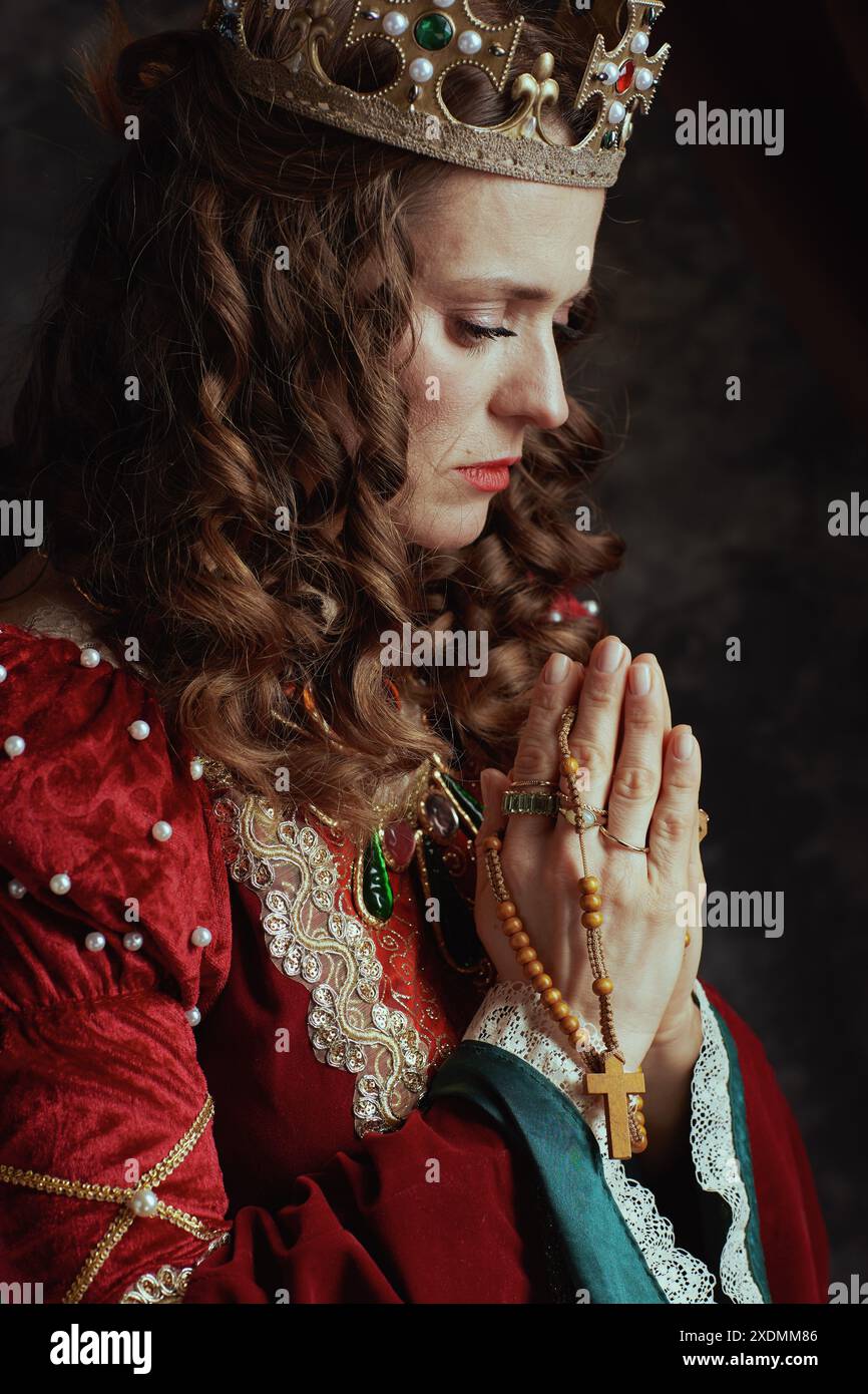 medieval queen in red dress with rosary and crown praying on dark gray ...