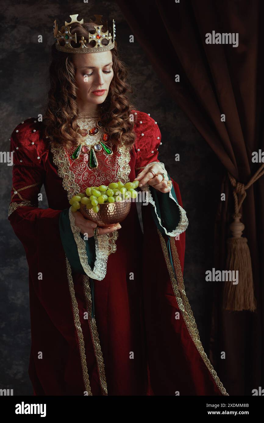 medieval queen in red dress with plate of grapes and crown on dark gray ...