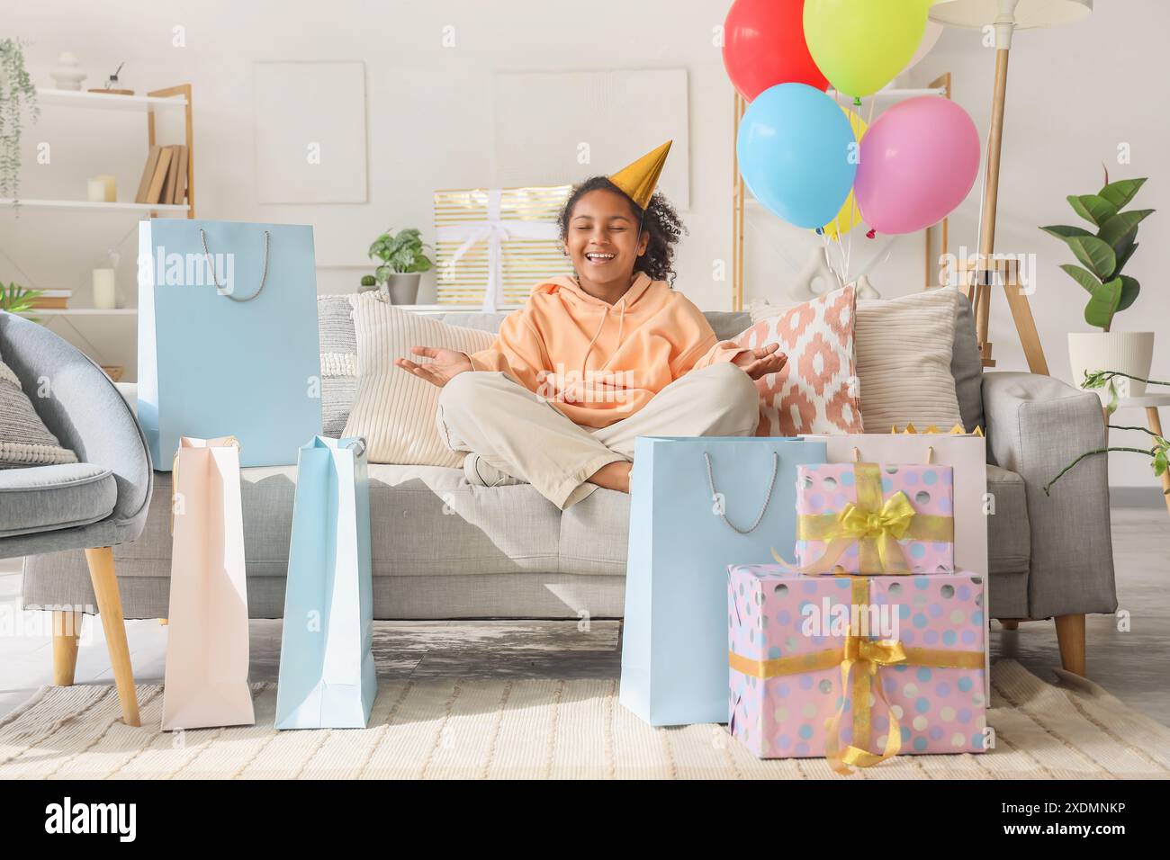 Happy African-American girl with gifts and balloons sitting on sofa at ...