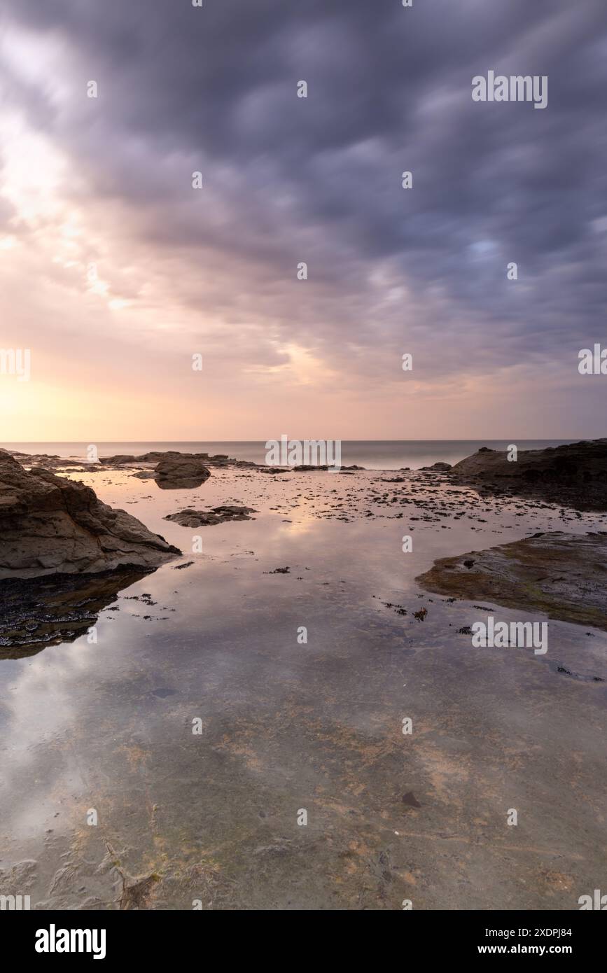 Rocky shoreline under a dramatic sunset sky reflecting on wet sand ...
