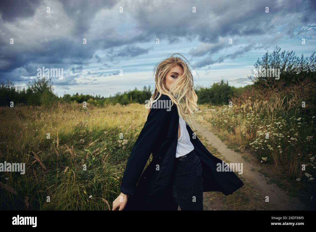 A woman walks down a dusty path through a field with her hair blowing ...