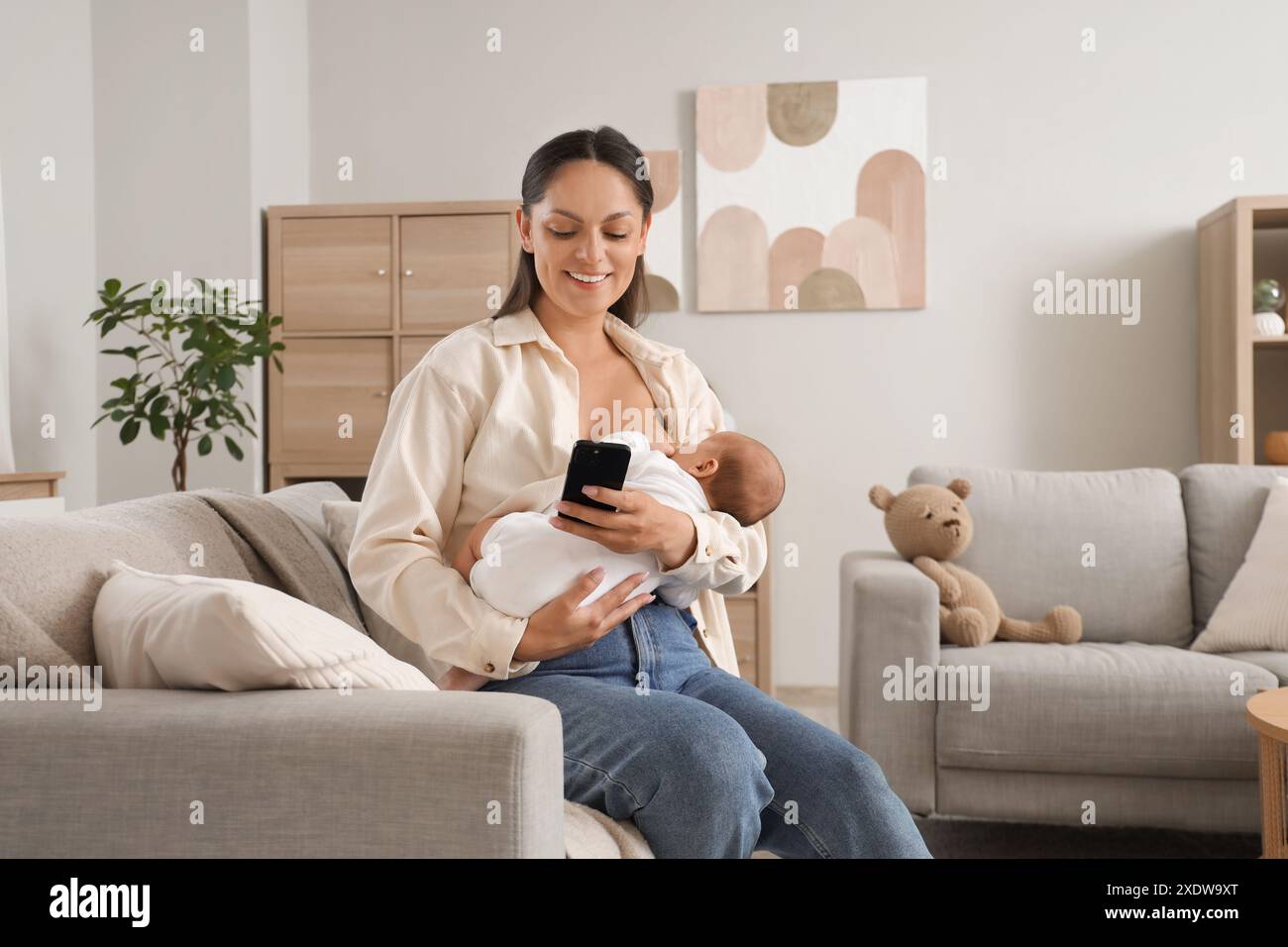 Mother breastfeeding her baby while using mobile phone on sofa at home ...