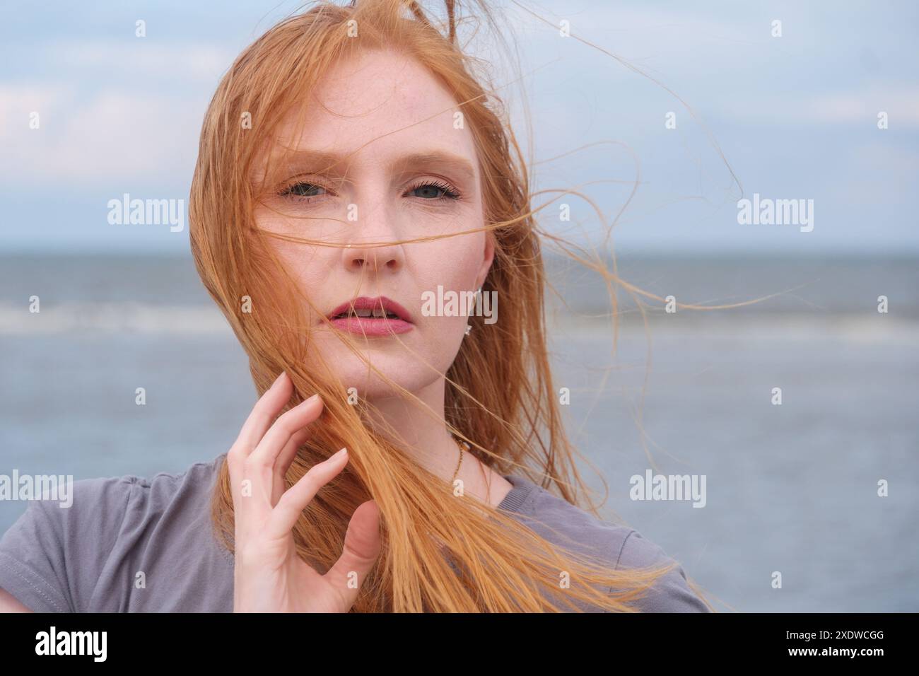 Woman with red hair stands by the sea, hair blowing across her face ...