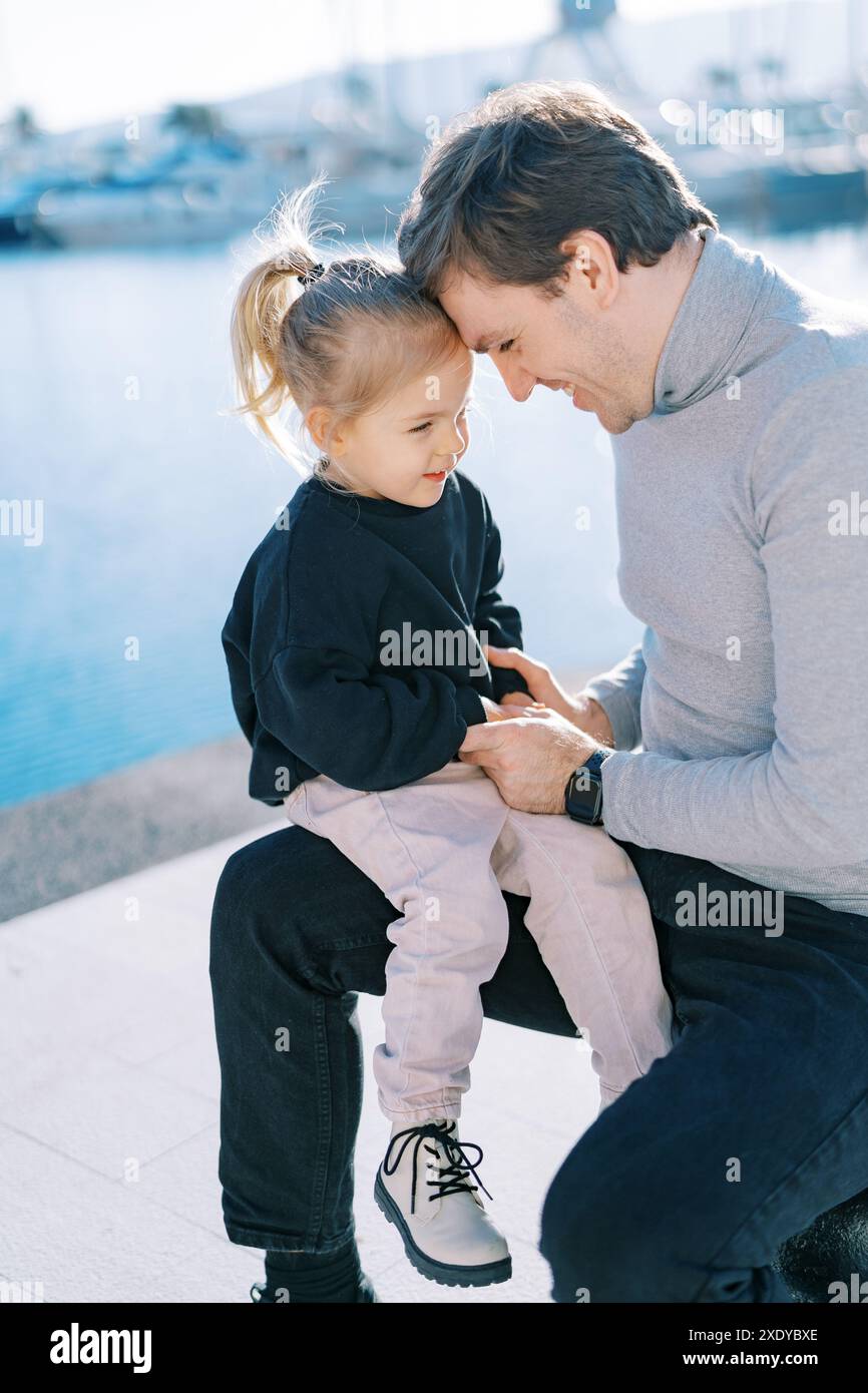 Smiling dad touching foreheads with little girl sitting on his knee, sitting on a bollard on the ...