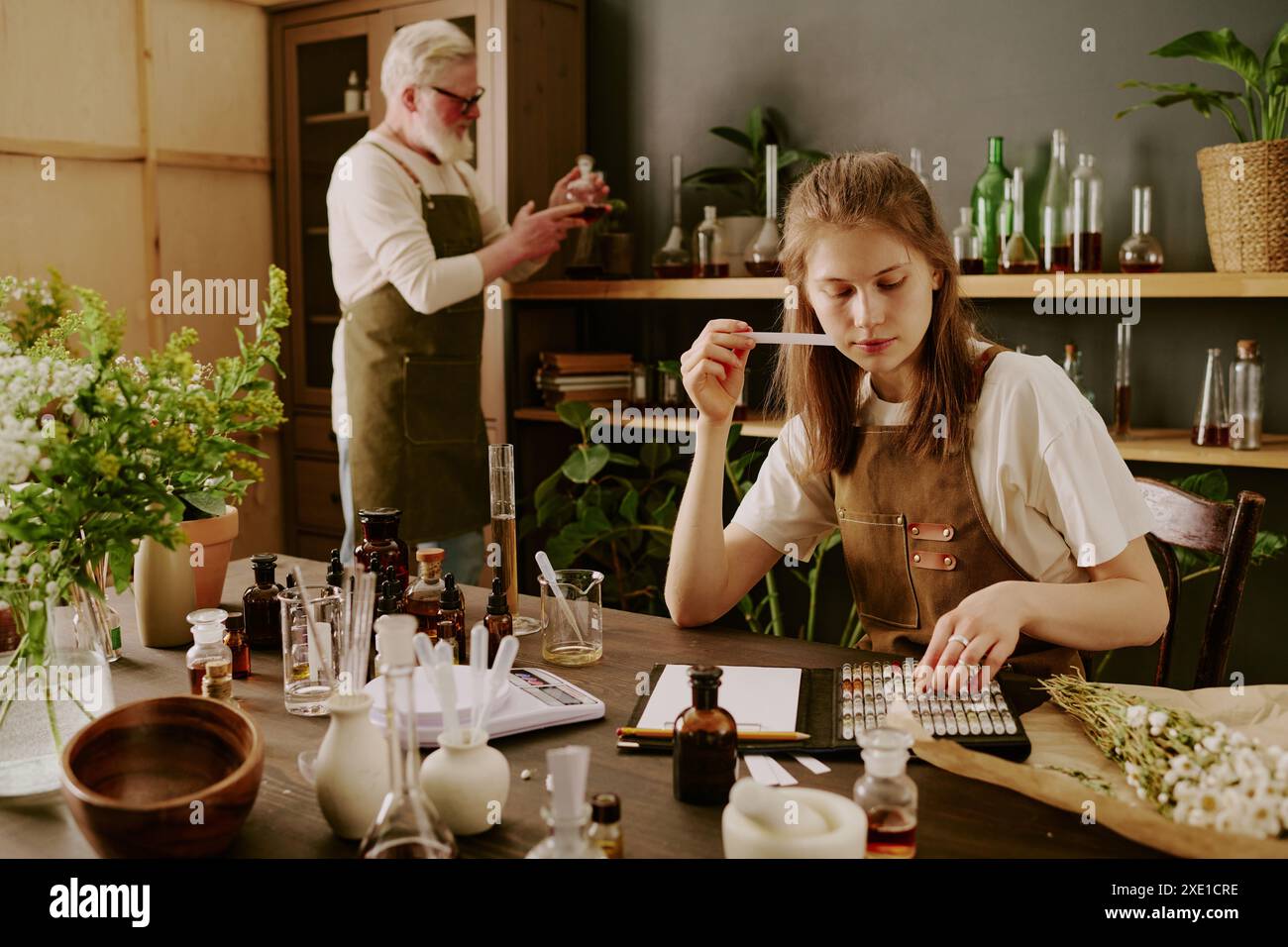 Female perfumer sitting at table and smelling tester while mature male ...