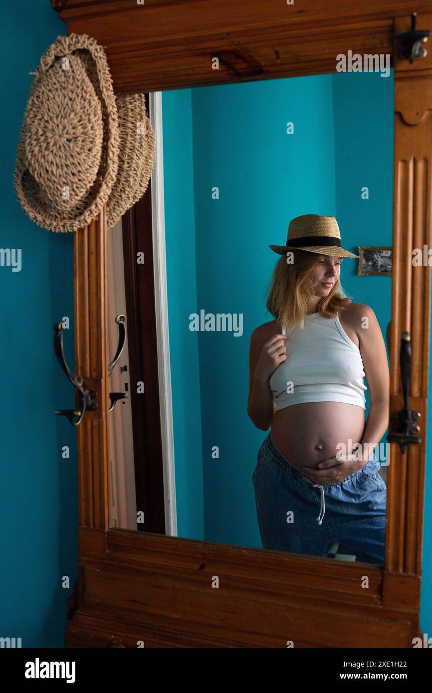Pregnant woman in hat, smiling at her reflection in a mirror, blue wall ...