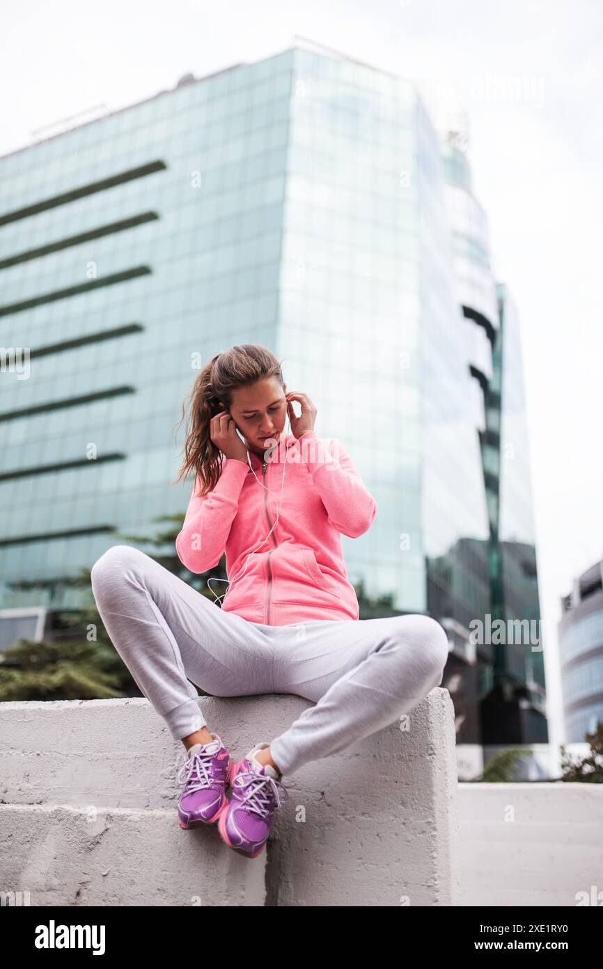 Fitness-focused young female relaxes on a ledge with her headphones on ...