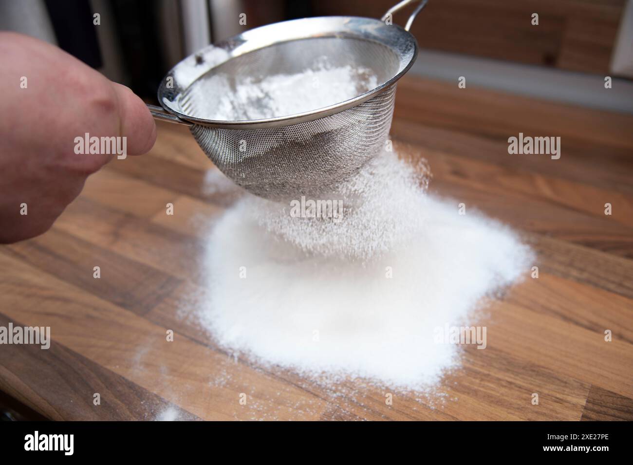 A close-up of a male hand sifting powdered flour onto a wooden surface ...