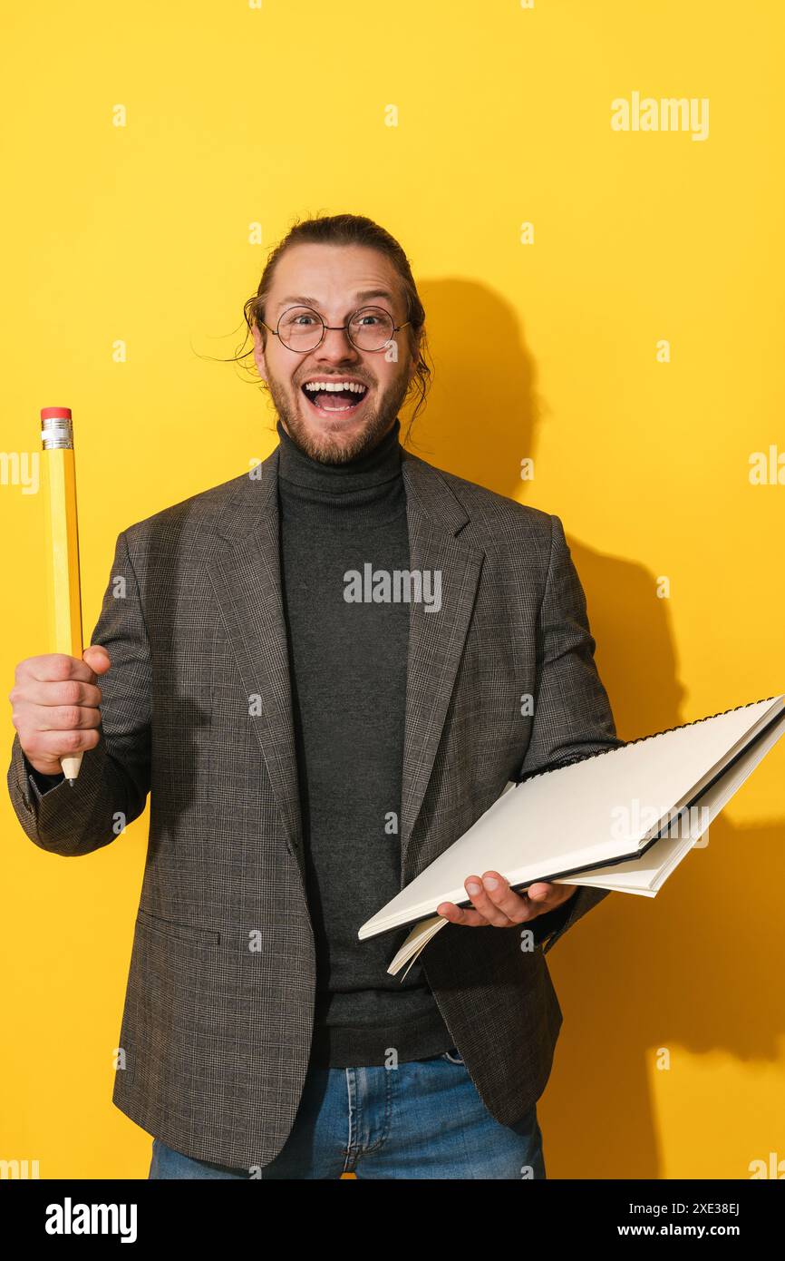 Cheerful man wearing glasses holding big pencil and notebook on yellow ...