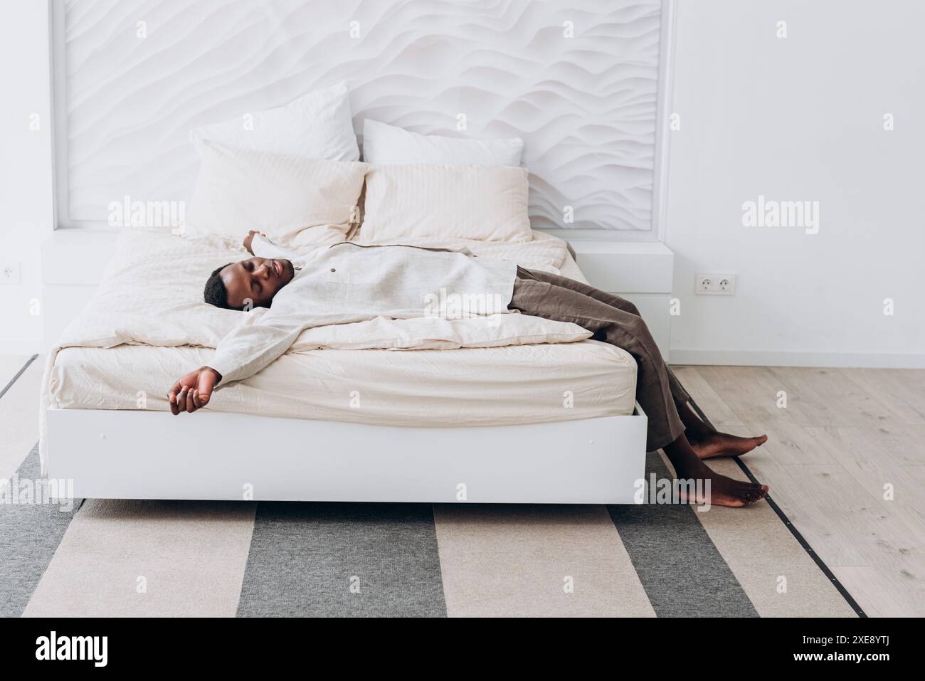 African American man lies spread out on neatly made bed in modern bedroom, enjoying moment of ...