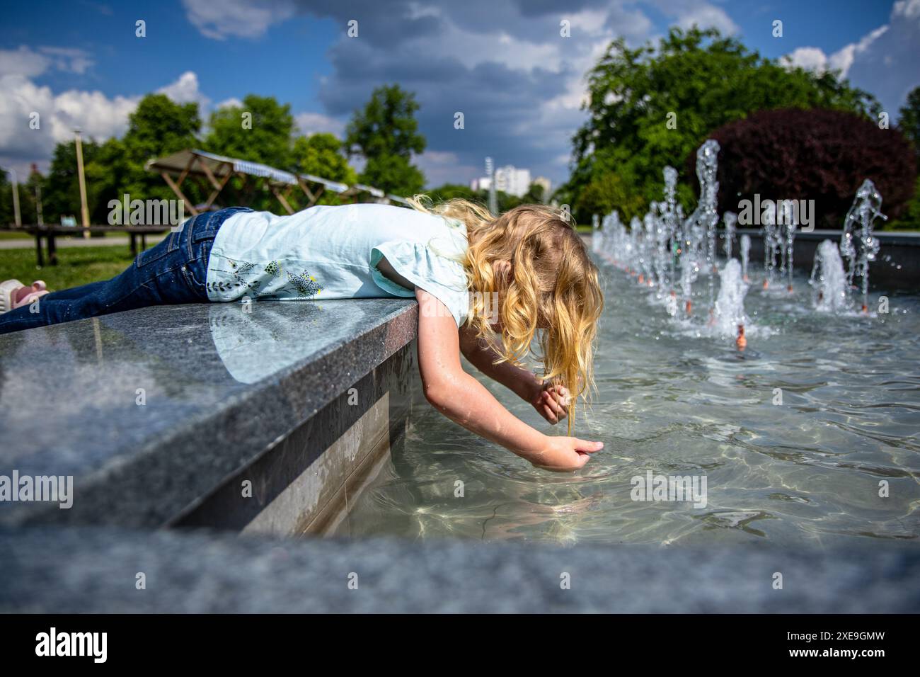 little girl in jeans and a light blue shirt splashes water from a city ...