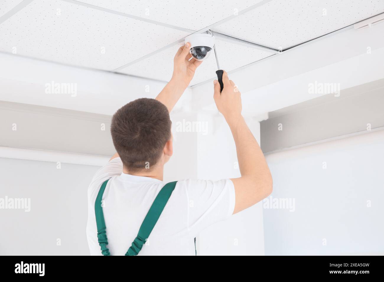 Technician with screwdriver installing CCTV camera on ceiling indoors ...
