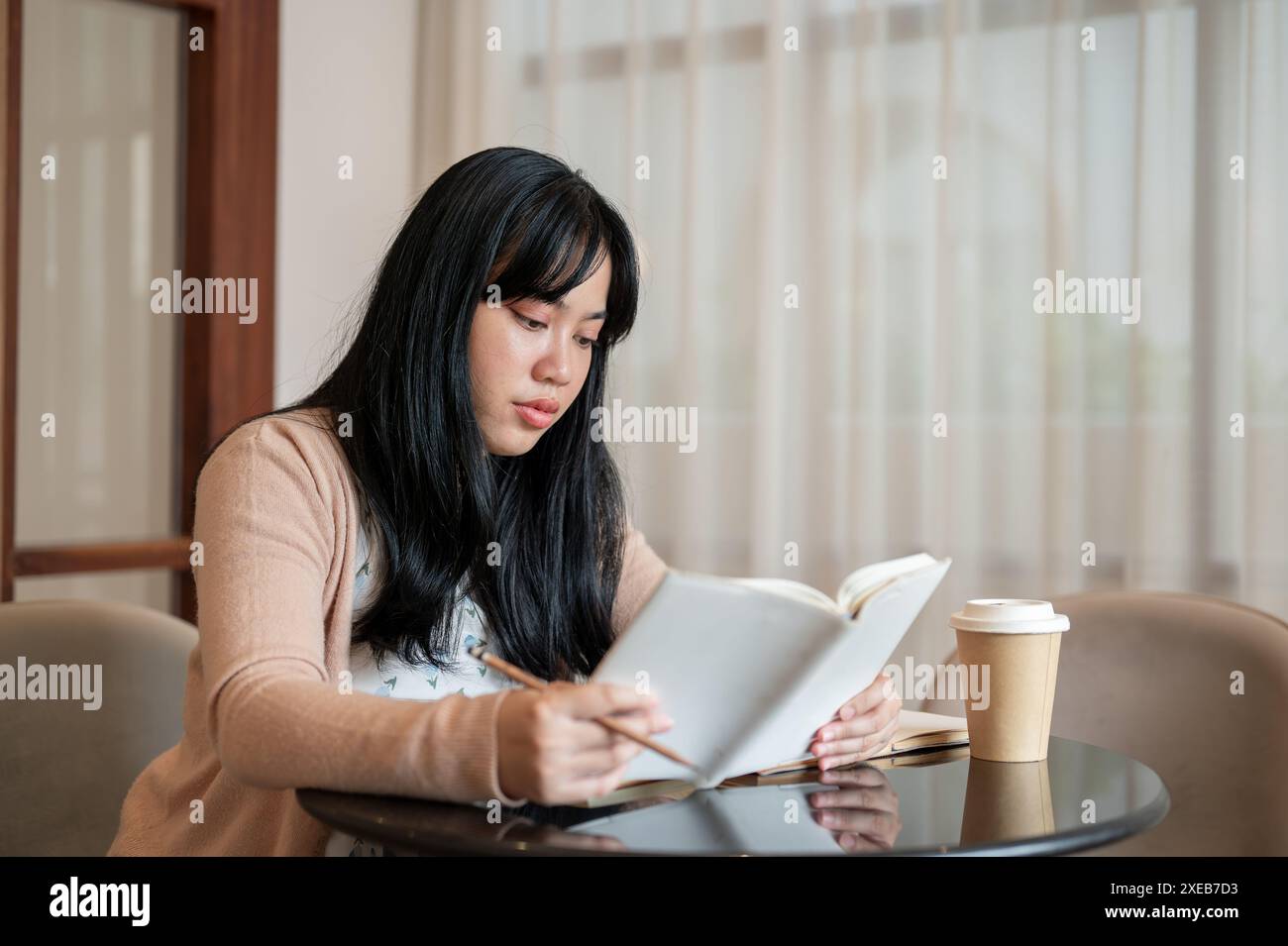 A young Asian woman focusing on reading a book at a table in a coffee ...
