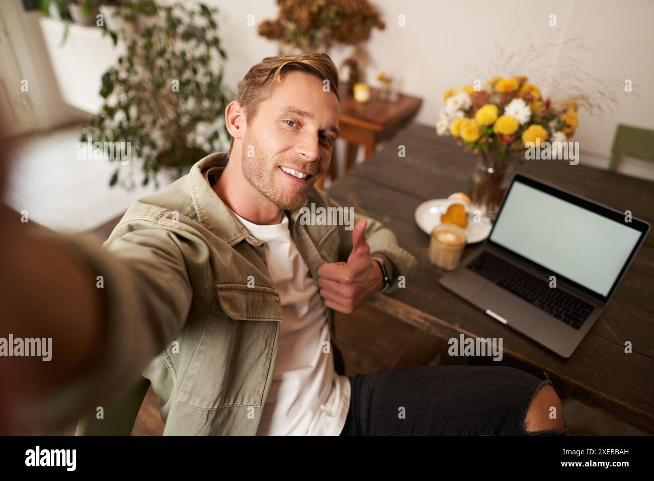 Man taking selfie with thumbs up, sitting in cafe with his laptop and ...