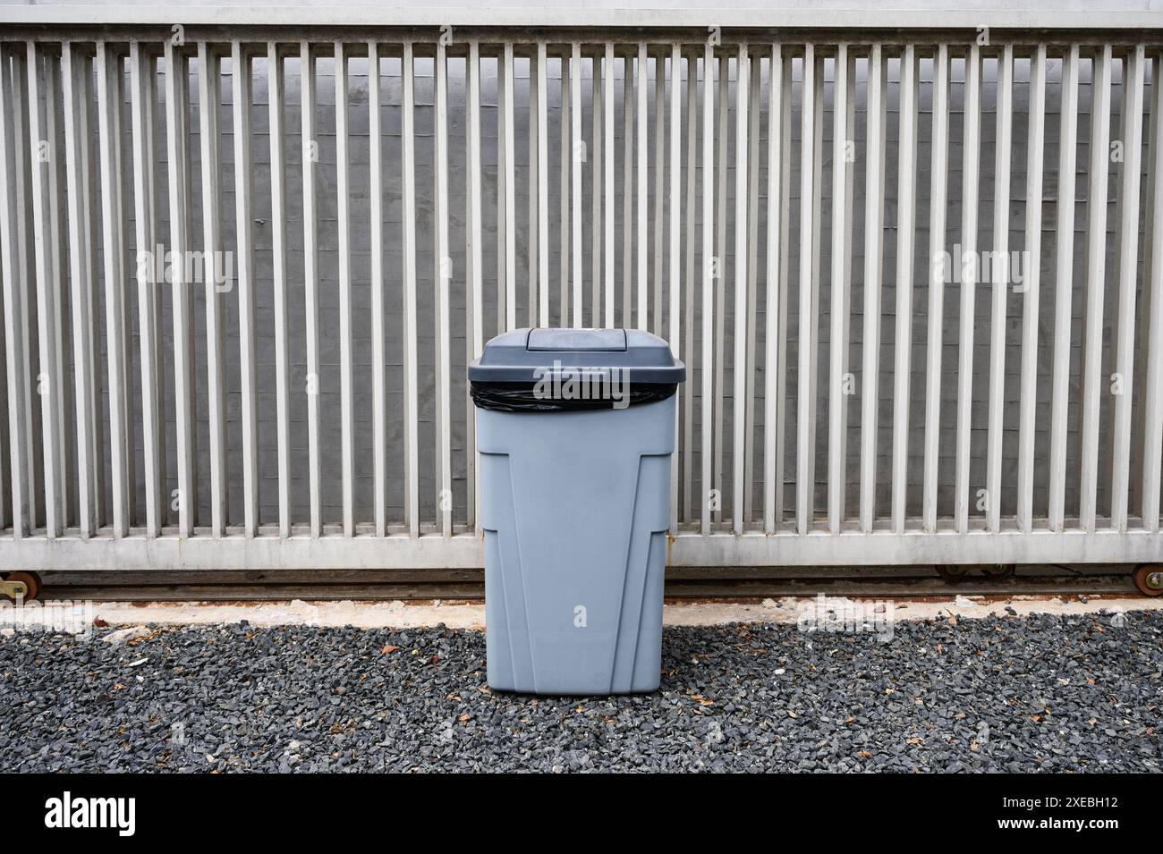 Gray garbage bin with plastic bag at metallic fence on gravel floor ...