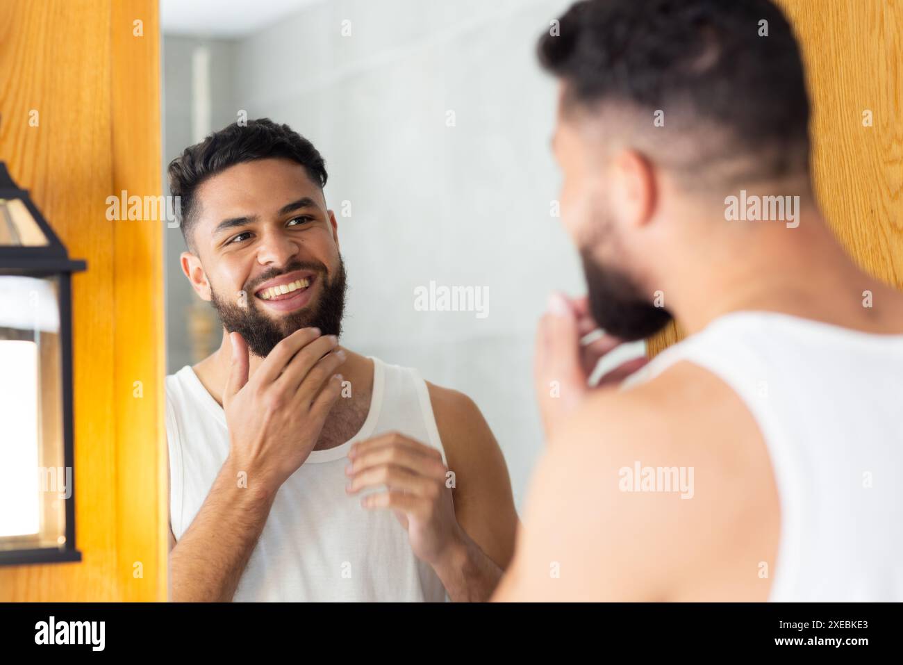 Man grooming beard and smiling in mirror, enjoying self-care at home ...