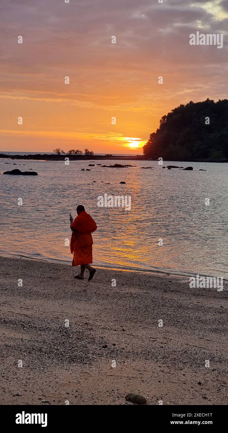 A lone Thai monk figure strolls along the sandy beach as the sun sets ...