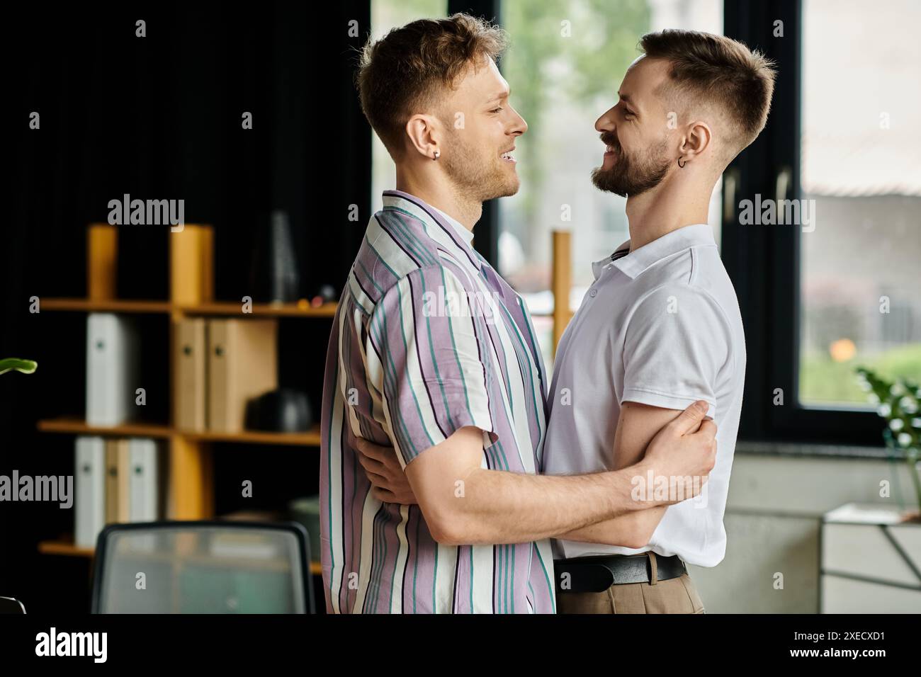 Two men, in casual attire, stand together in an office setting, smiling ...