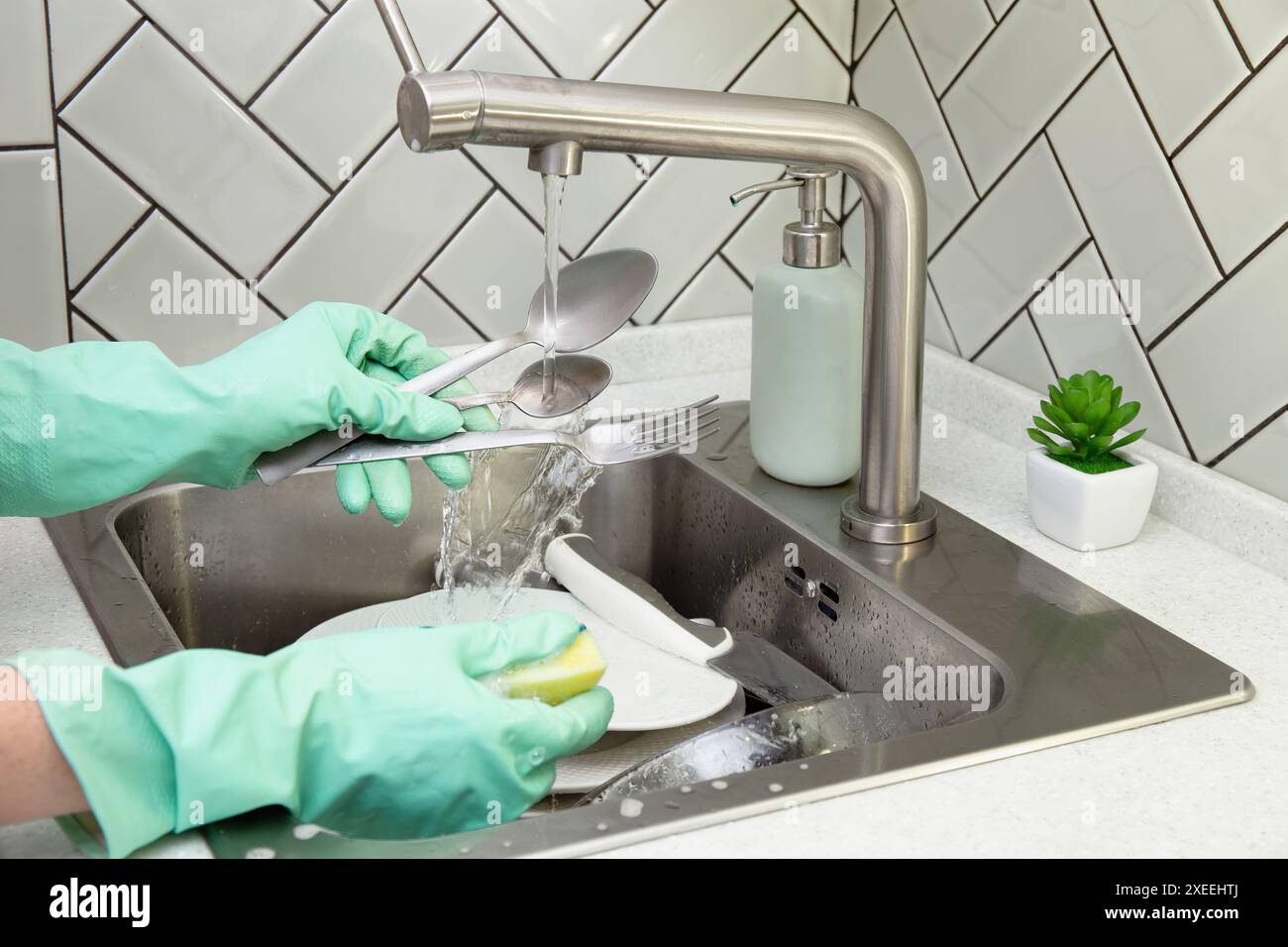 Hands wearing green gloves washing dishes and utensils in a stainless ...