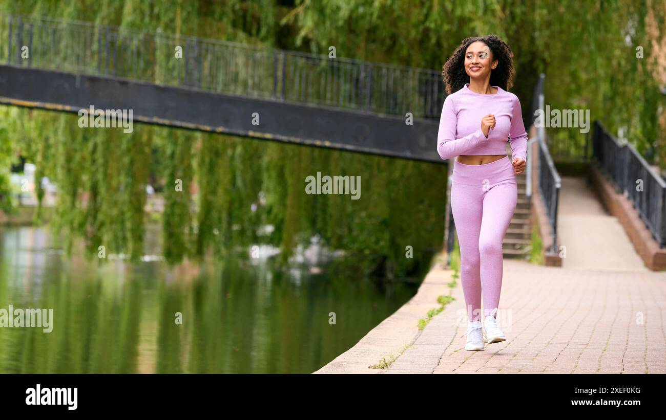 Woman Wearing Fitness Clothing Running Along Path By River In Urban ...