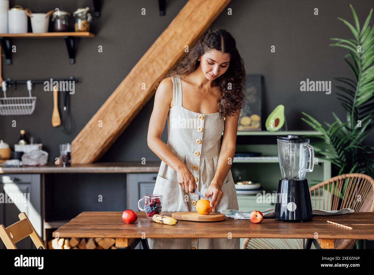 Charming curly-haired brunette girl in linen sundress prepares fruit ...