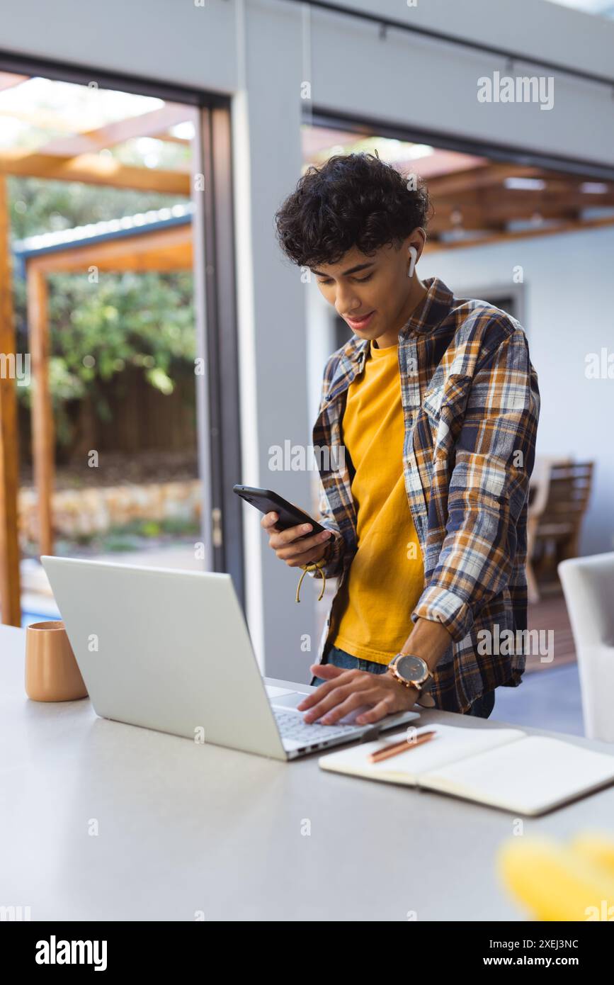 Using smartphone and laptop, teenage boy working from home in kitchen. Remote working ...