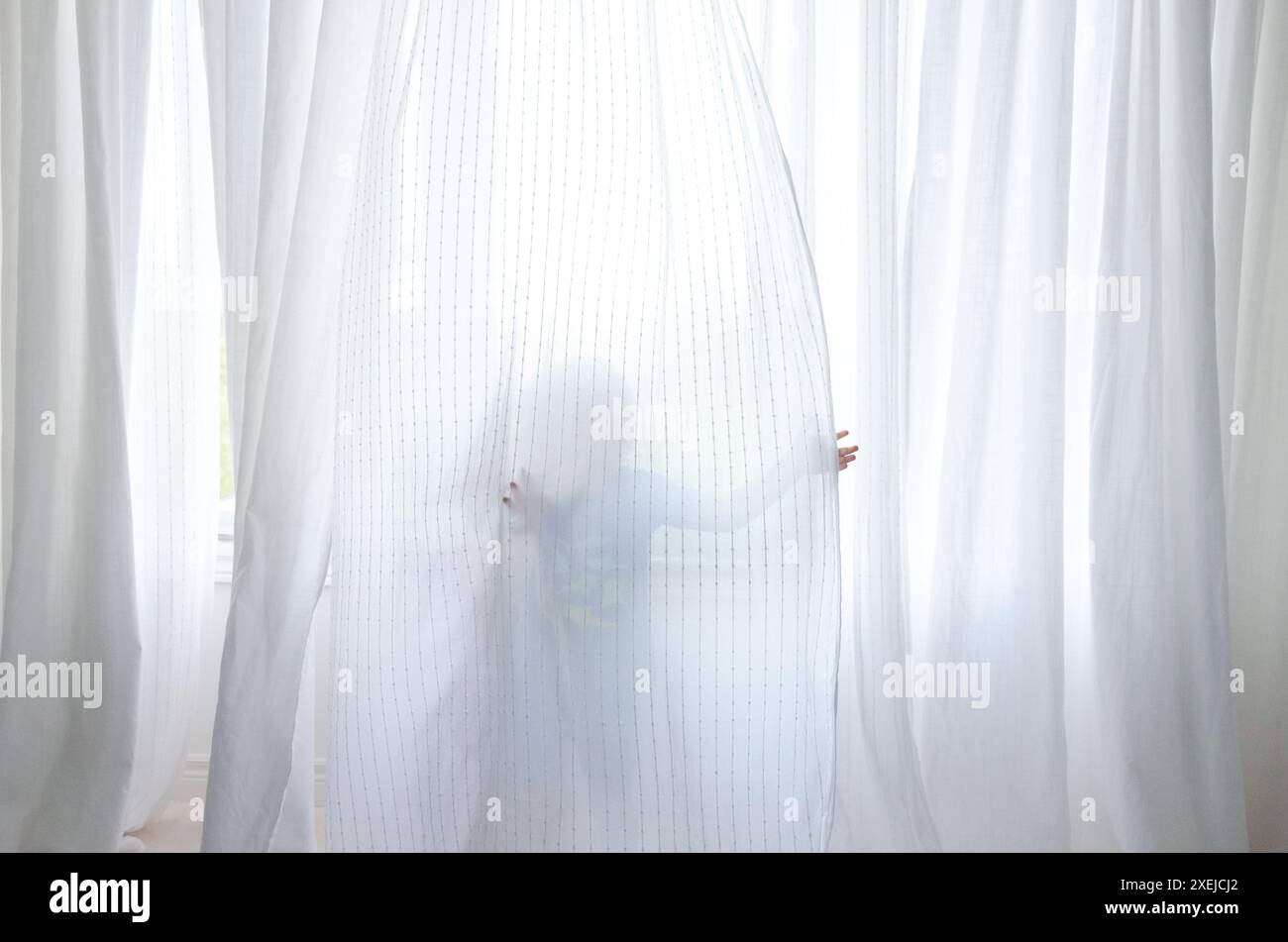 A child playing behind sheer white curtains, partially visible fabric ...