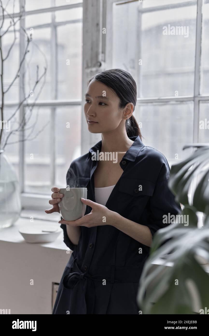 Asian female ceramic artist standing near window with cup Stock Photo ...