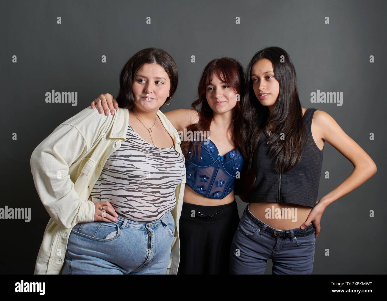 Three teenage girls stand side by side against a gray background. The ...