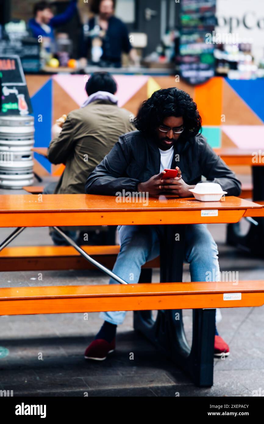 A man sits at a bench table in a food hall checking his mobile phone ...