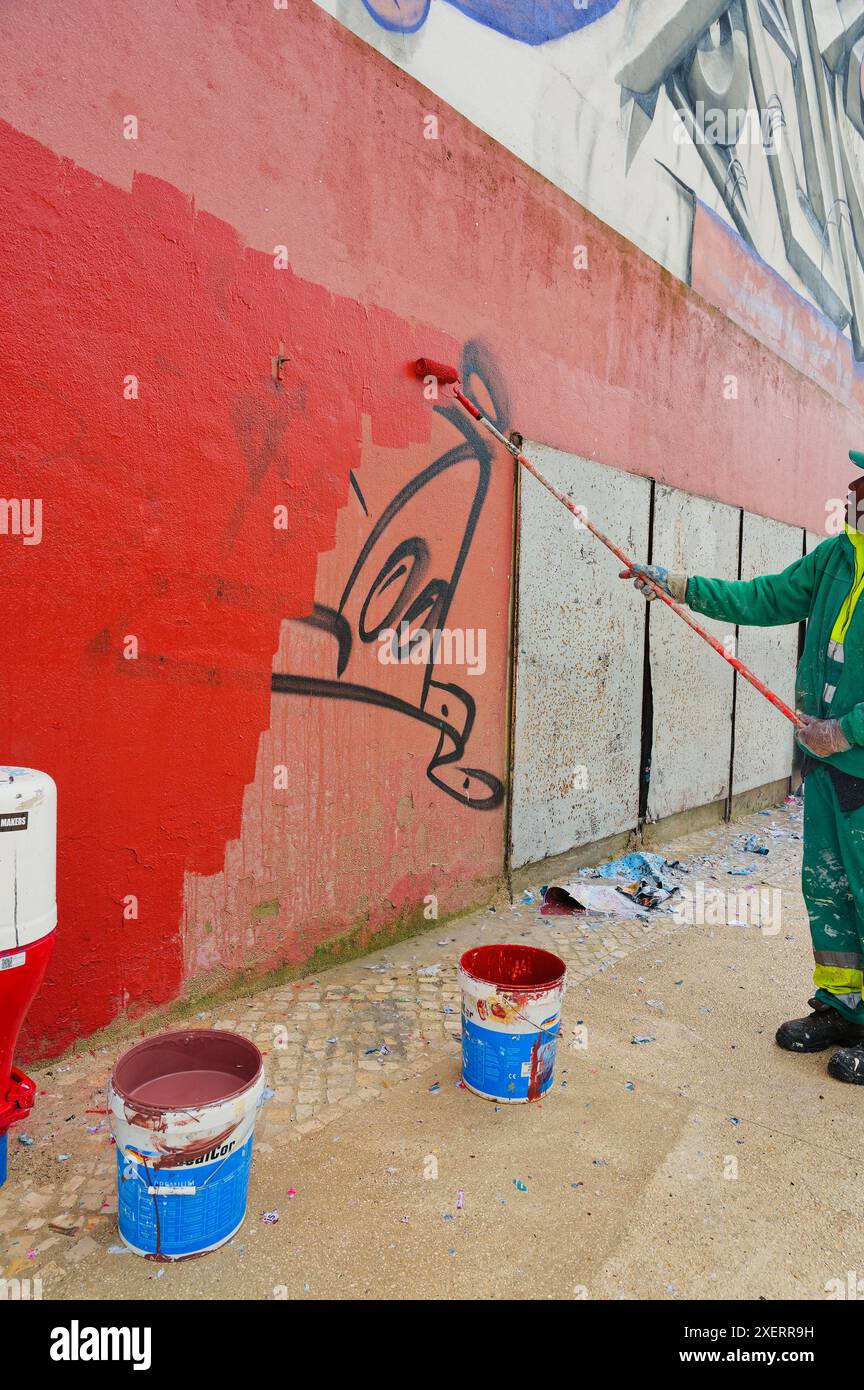 Process of a painter in green coveralls as he paints over graffiti on a red wall, covering old ...