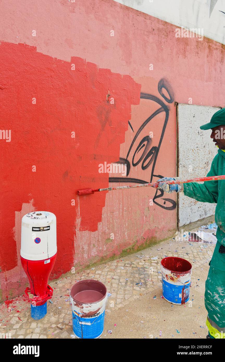 Process of a painter in green coveralls as he paints over graffiti on a red wall, covering old ...