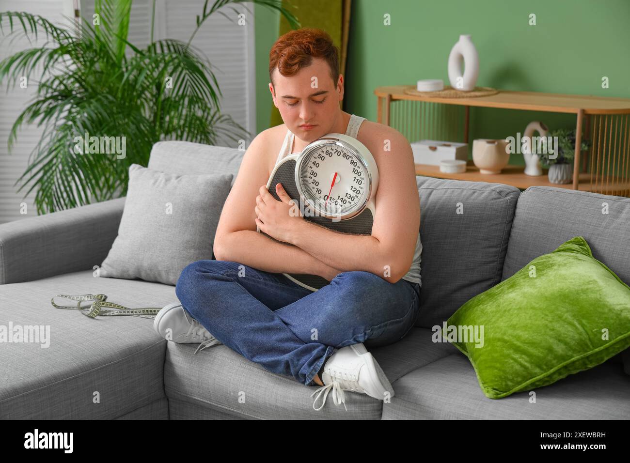 Young sad overweight man with scales sitting on sofa at home. Weight ...