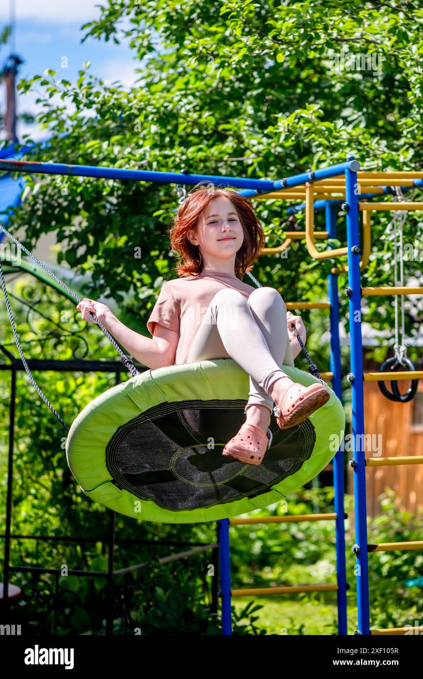 Cute beautiful teenage girl with red hair swings on a swing in summer countryside, vacation ...