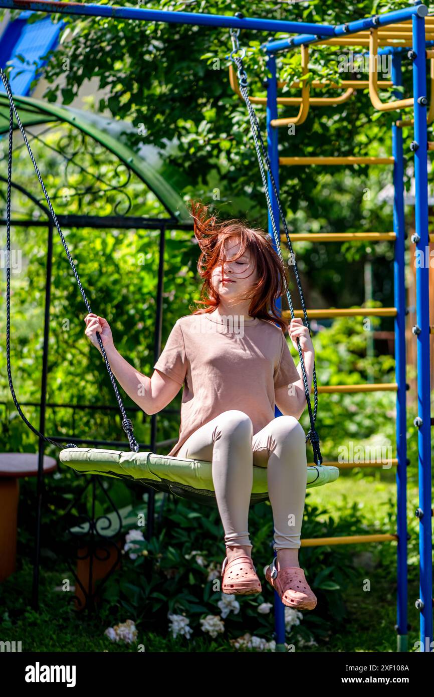 Cute beautiful teenage girl with red hair swings on a swing in summer countryside, vacation ...