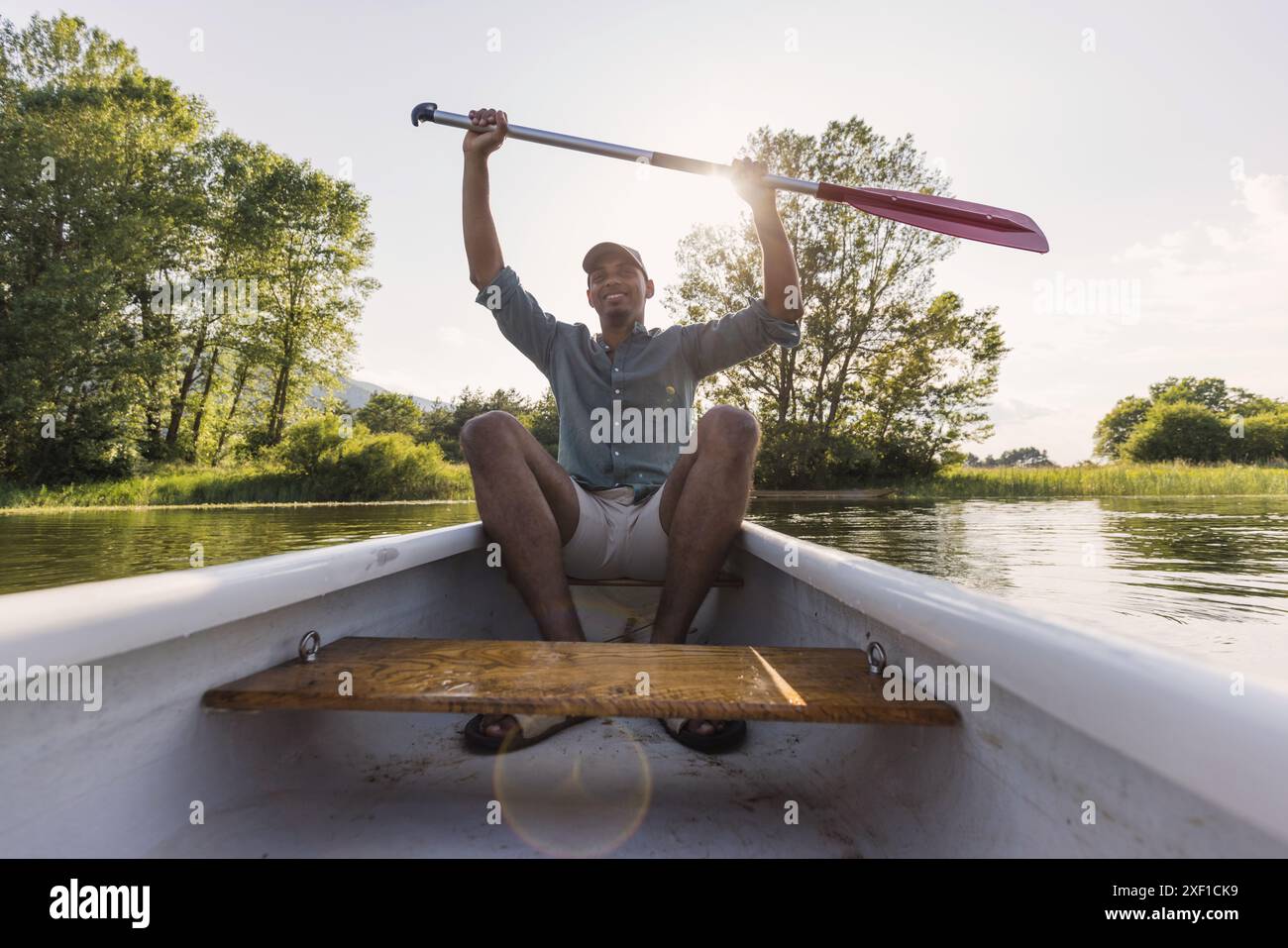 Cheerful young man sitting in a canoe on a calm lake and lifting a ...