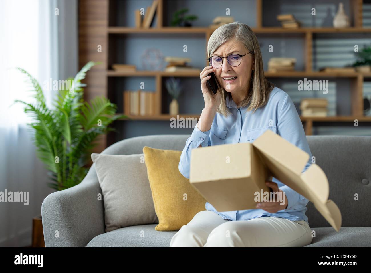 Elderly woman sitting on a couch, talking on a mobile phone, and opening a package. She looks ...