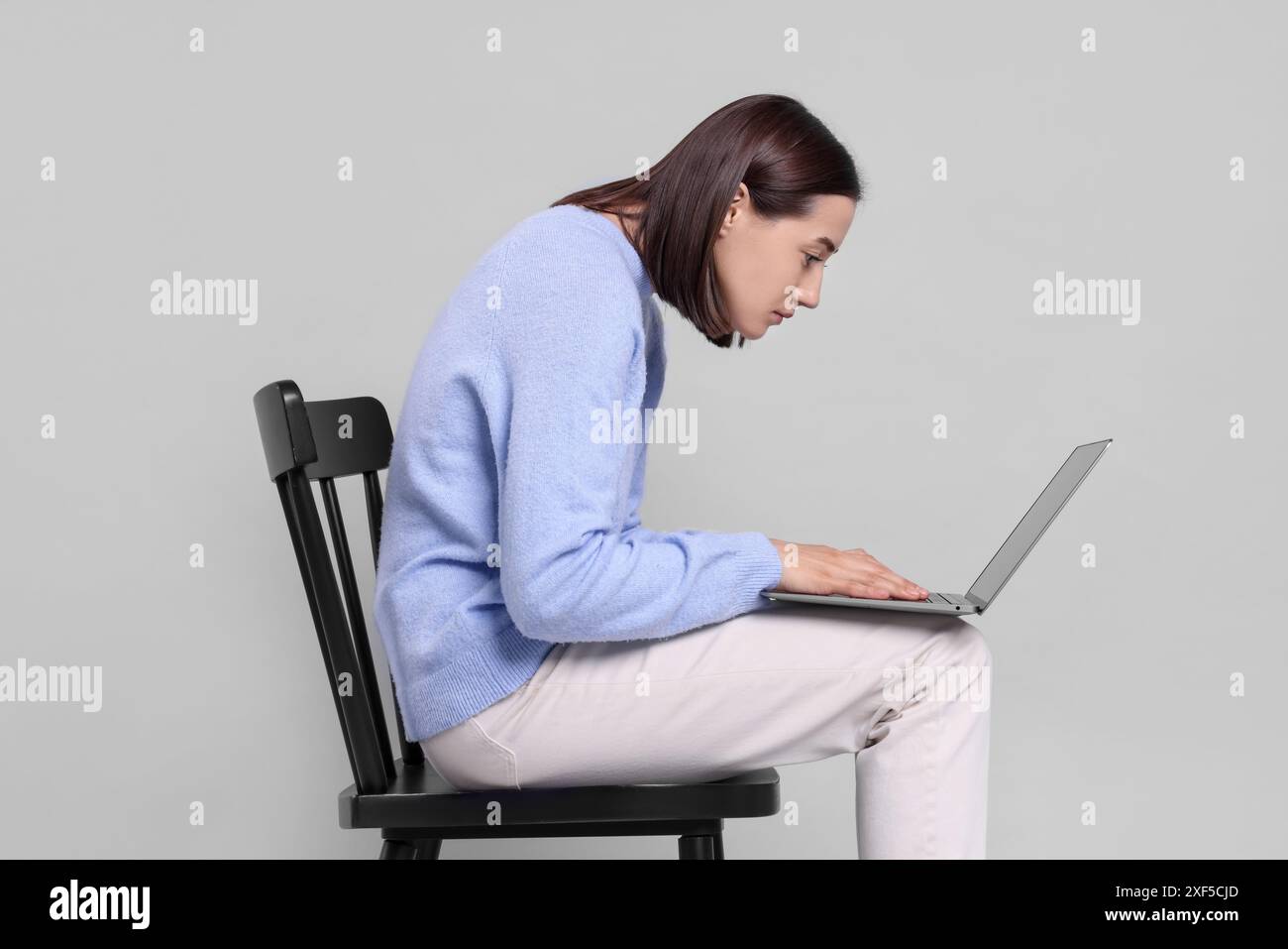 Woman with poor posture sitting on chair and using laptop against gray ...