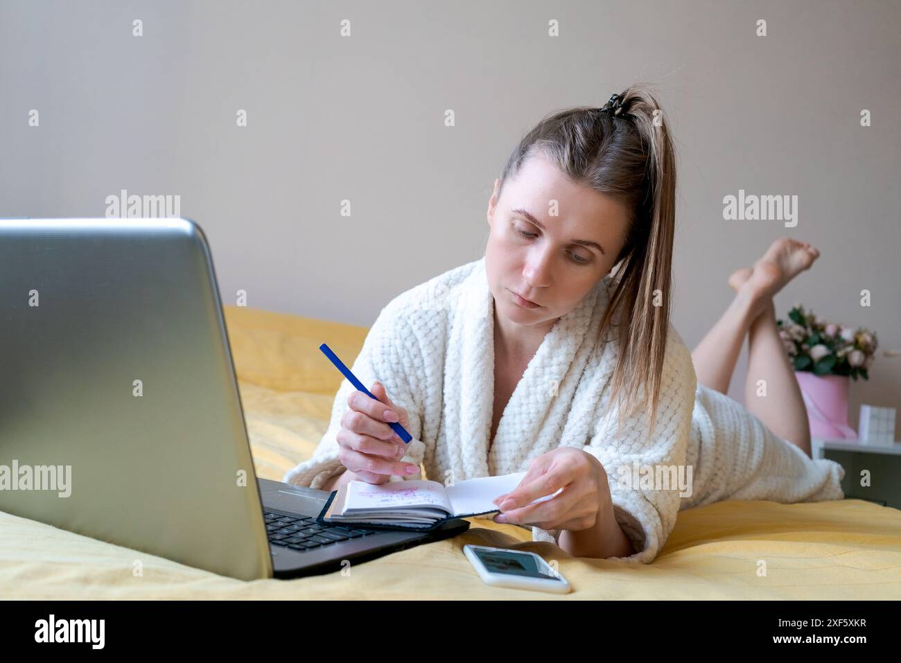 A woman is writing on a laptop while laying on a bed. She is wearing a ...