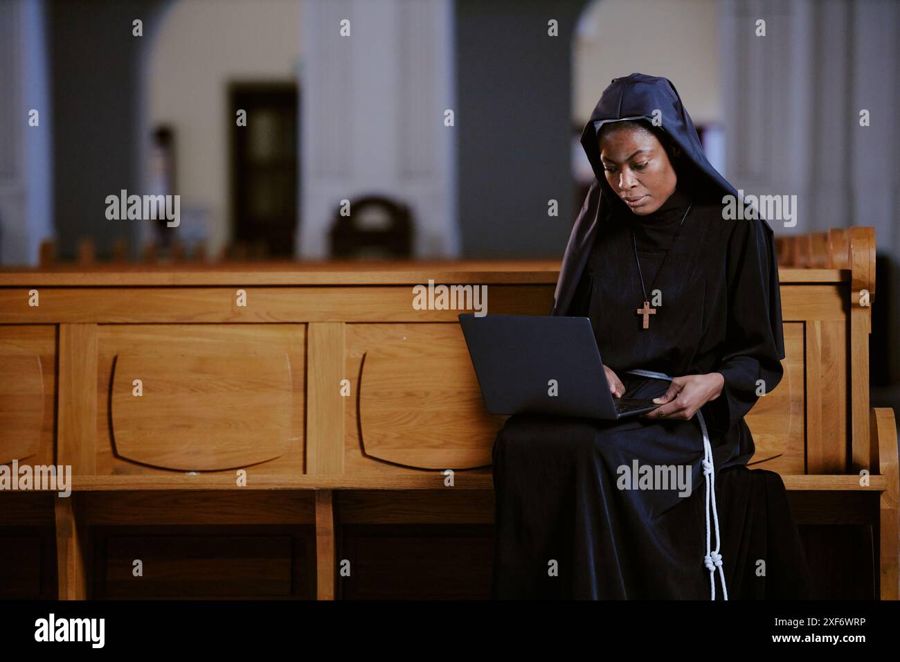 Medium shot of young African American nun sitting on wooden pew typing ...