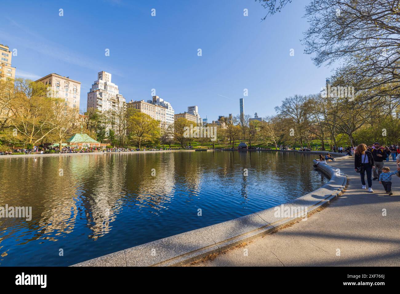 Pedestrians stroll along lakeside paths in Central Park, framed by towering skyscrapers of ...