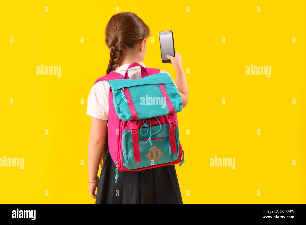 Little schoolgirl with backpack and mobile phone on yellow background ...