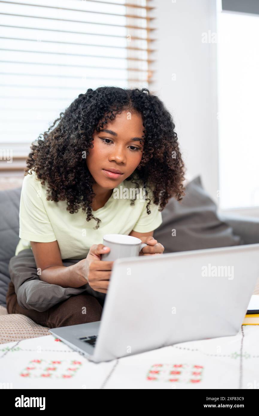 An attractive young Black woman is having coffee while reading online articles on her laptop ...