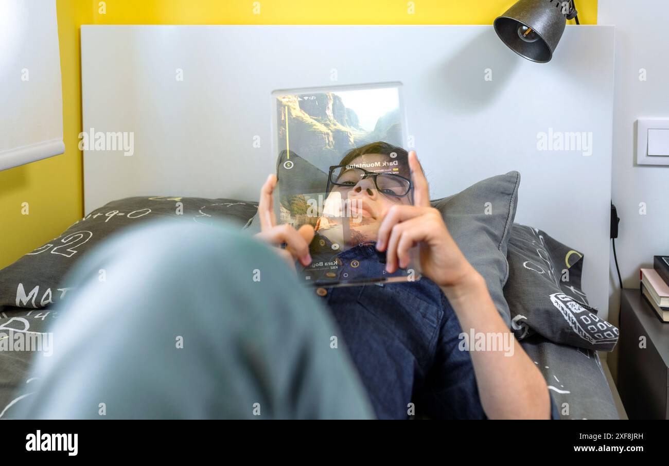 Relaxed boy watching video in a futuristic transparent tablet laying over his bed. Digital ...