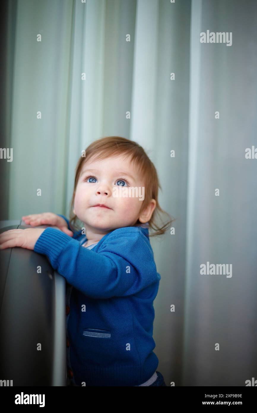 A baby with blue eyes and a blue sweater looks up with a curious ...