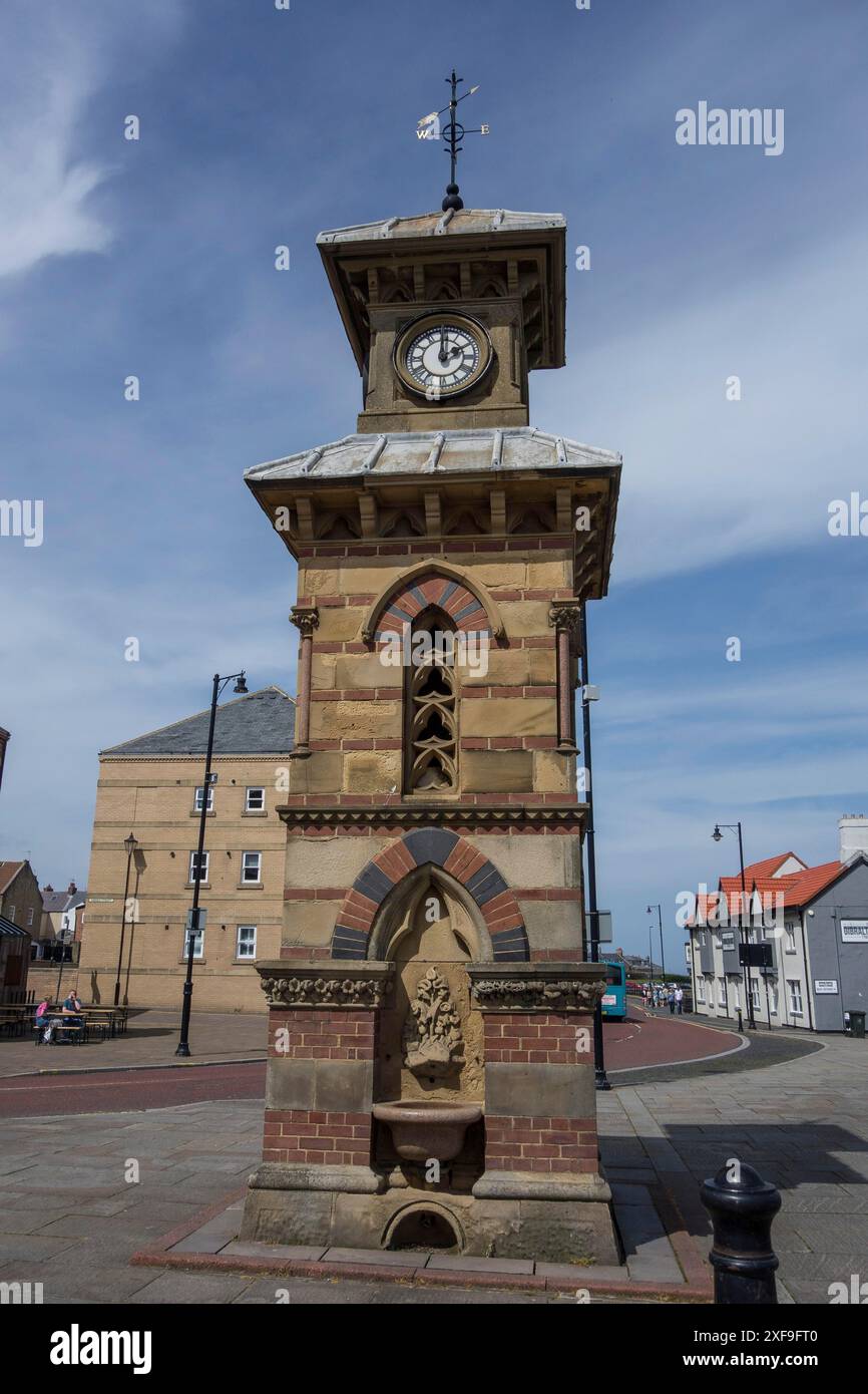 A brick clock tower with a fountain stands on a street under a cloudy ...