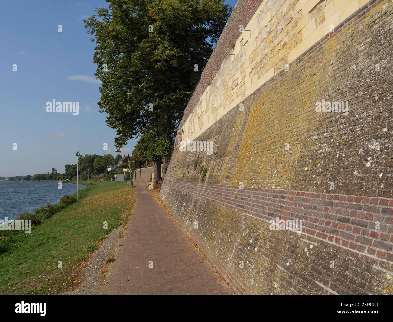 A paved path along a high brick wall and a river, flanked by trees, in ...