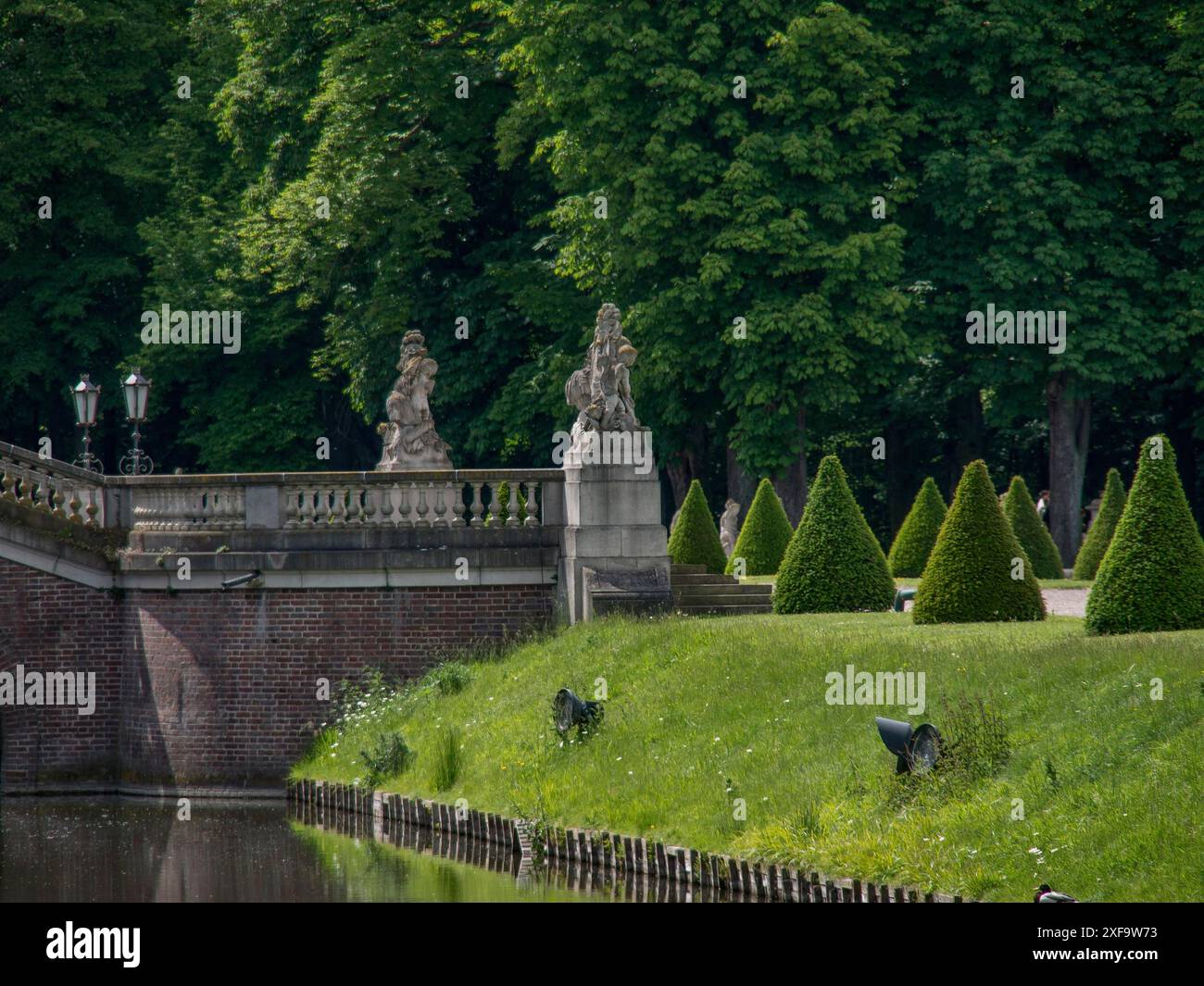 A stone bridge with statues leads over a river, flanked by neat hedges ...