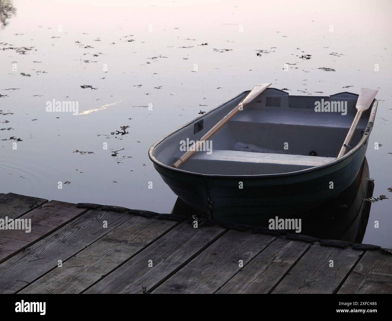 A green rowing boat lies on a wooden jetty on calm water at dusk ...