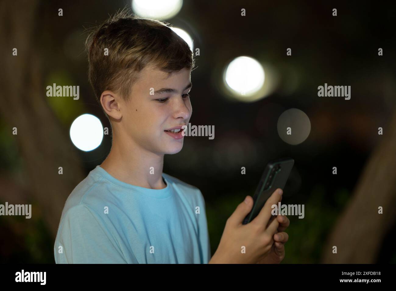 Teenage boy texting with his friend on cellphone outdoors on night city ...